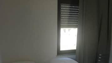 Quarto de hotel com duas camas de solteiro, cada uma com colcha azul e almofadas brancas. Há uma janela com persianas, uma mesinha de cabeceira de madeira e uma cadeira.