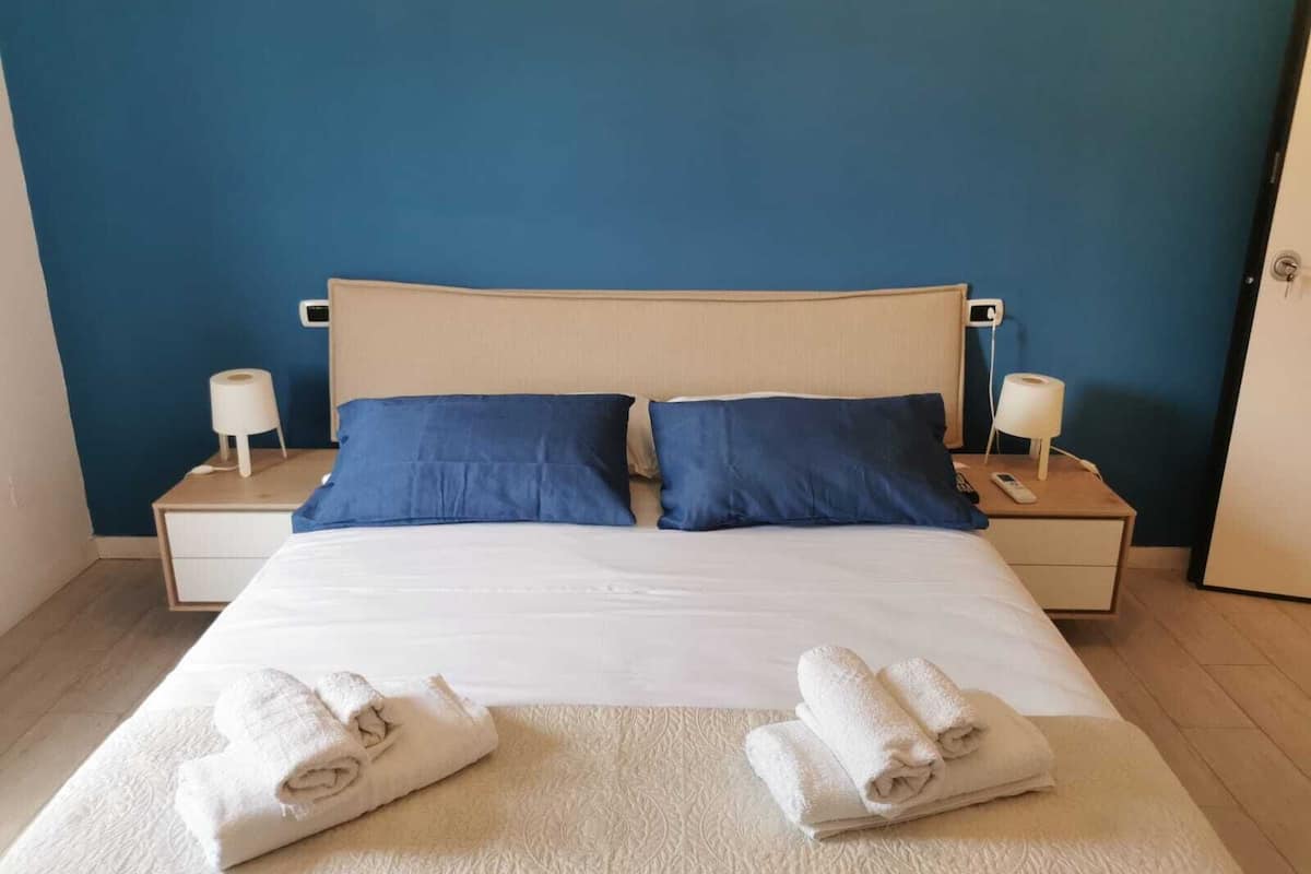 Cama bem arrumada com duas almofadas, duas mesas de cabeceira com luminárias e dois rolinhos de toalha sobre um edredom texturizado.