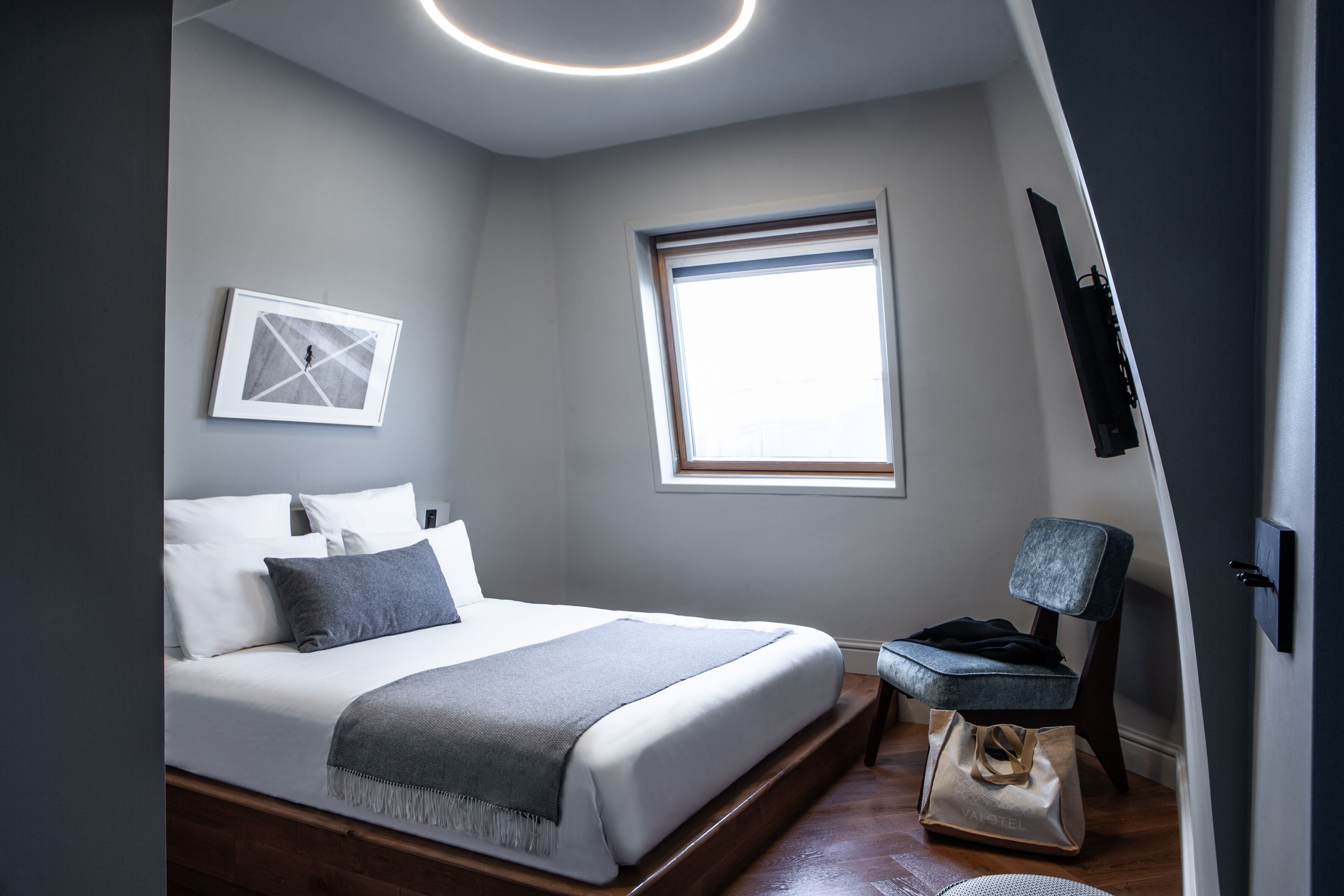 Quarto moderno com cama grande, janela, cadeira e TV fixada na parede.
