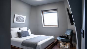 Quarto moderno com cama grande, janela, cadeira e TV fixada na parede.
