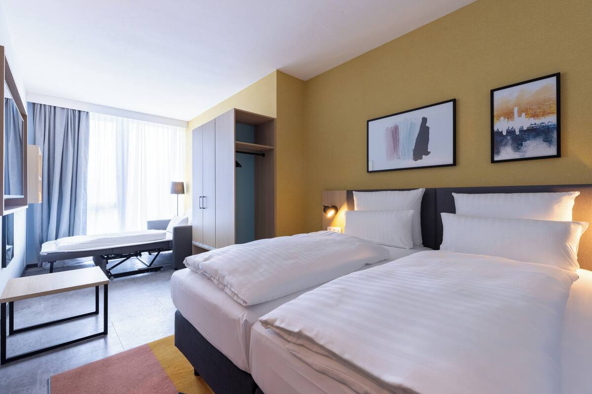 Quarto de hotel com duas camas, uma escrivaninha, uma cadeira, uma mesa de cabeceira e uma janela com cortinas.