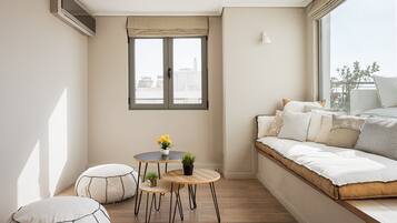 Um quarto pequeno e acolhedor com um banco embutido, duas mesas redondas e uma janela com vista para edifícios.