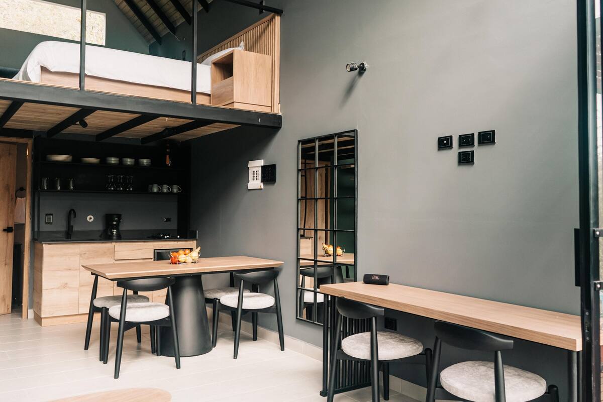 Quarto compacto e moderno com beliche, cozinha compacta, área de refeições com mesa e cadeiras de madeira, e um espelho grande na parede.