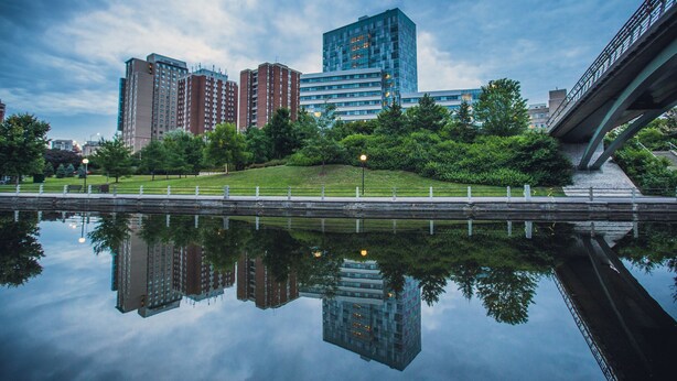 Paisagem urbana com edifícios modernos altos, um parque com um espelho d'água e uma ponte para pedestres.