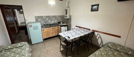 Uma cozinha compacta com geladeira, fogão, pia e armários de madeira. Há uma mesa de jantar com cadeiras e um ventilador de teto.