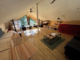 Um amplo loft com pé direito alto, piso de madeira, área de jantar com mesa e cadeiras, um espaço de estar aconchegante com sofá e uma grande janela com vista para o verde lá fora.