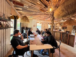 Um interior rústico com uma longa mesa comunitária, vigas de madeira no teto e um espaço de bar com prateleiras cheias de garrafas.