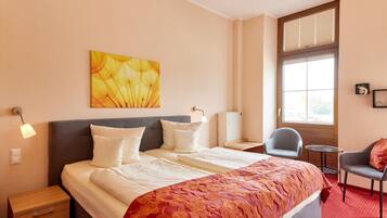 Quarto de hotel com uma cama grande, uma mesinha de cabeceira, uma cadeira, uma pequena mesa e uma janela com persianas.
