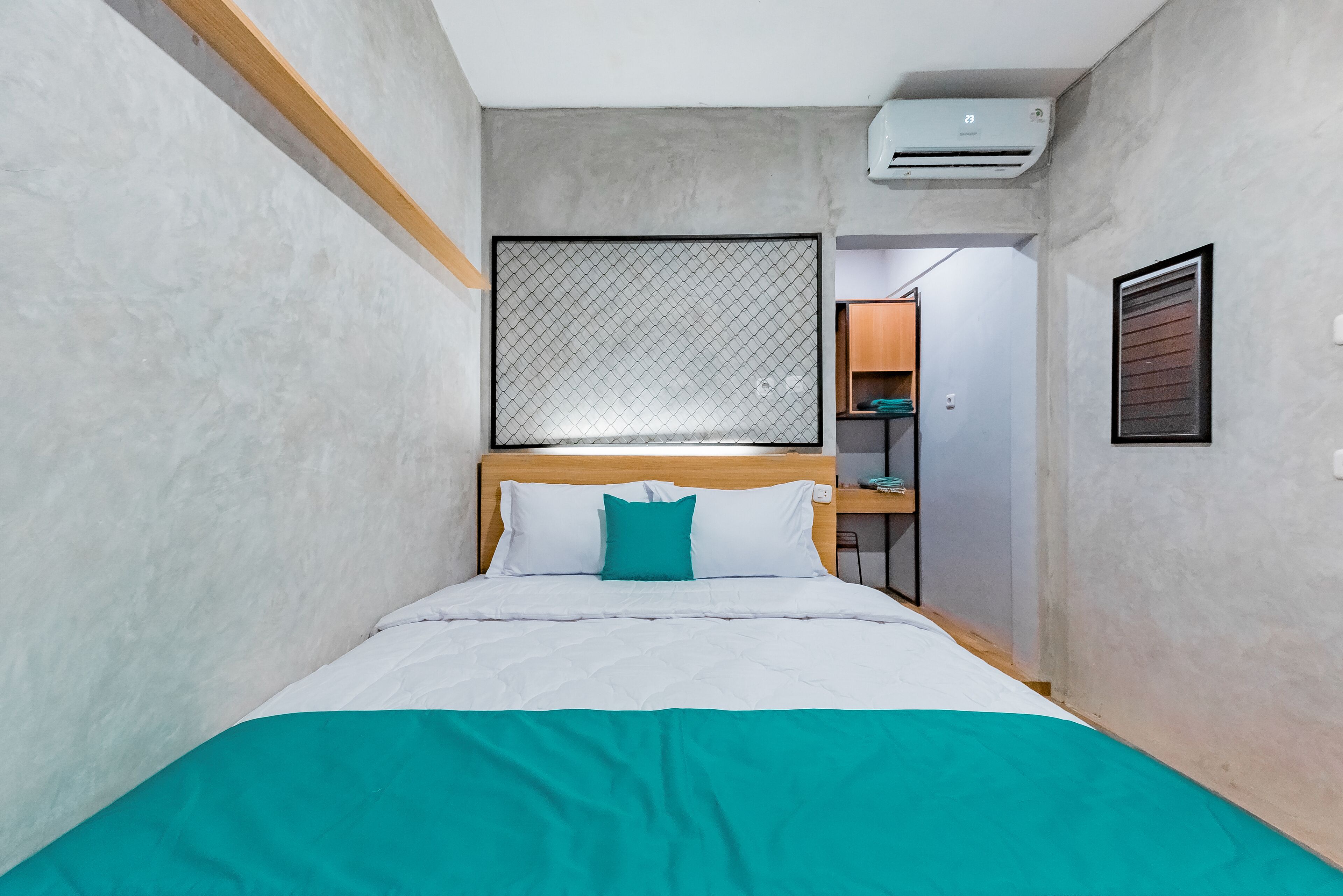 Quarto pequeno e estreito com uma cama, cabeceira, janela com persianas, ar condicionado fixado na parede e uma pequena escrivaninha com cadeira.
