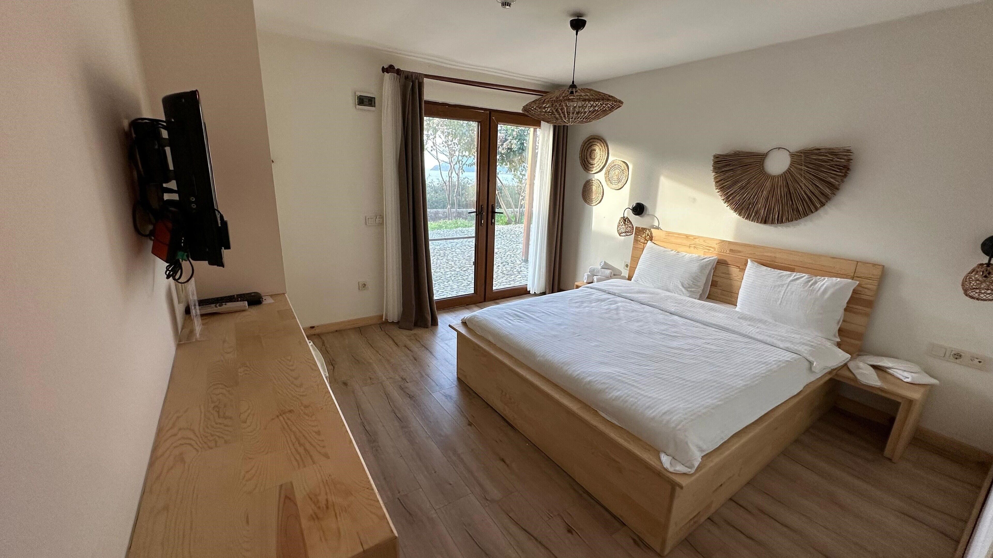 Quarto com cama de madeira, mesa de cabeceira, televisão na parede e vista através de uma porta de vidro.