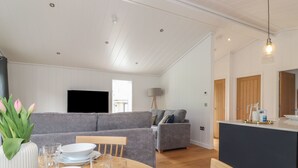 Uma sala de estar moderna com um sofá cinzento, uma mesa de jantar posta com pratos e copos, e uma televisão de tela plana fixada na parede.