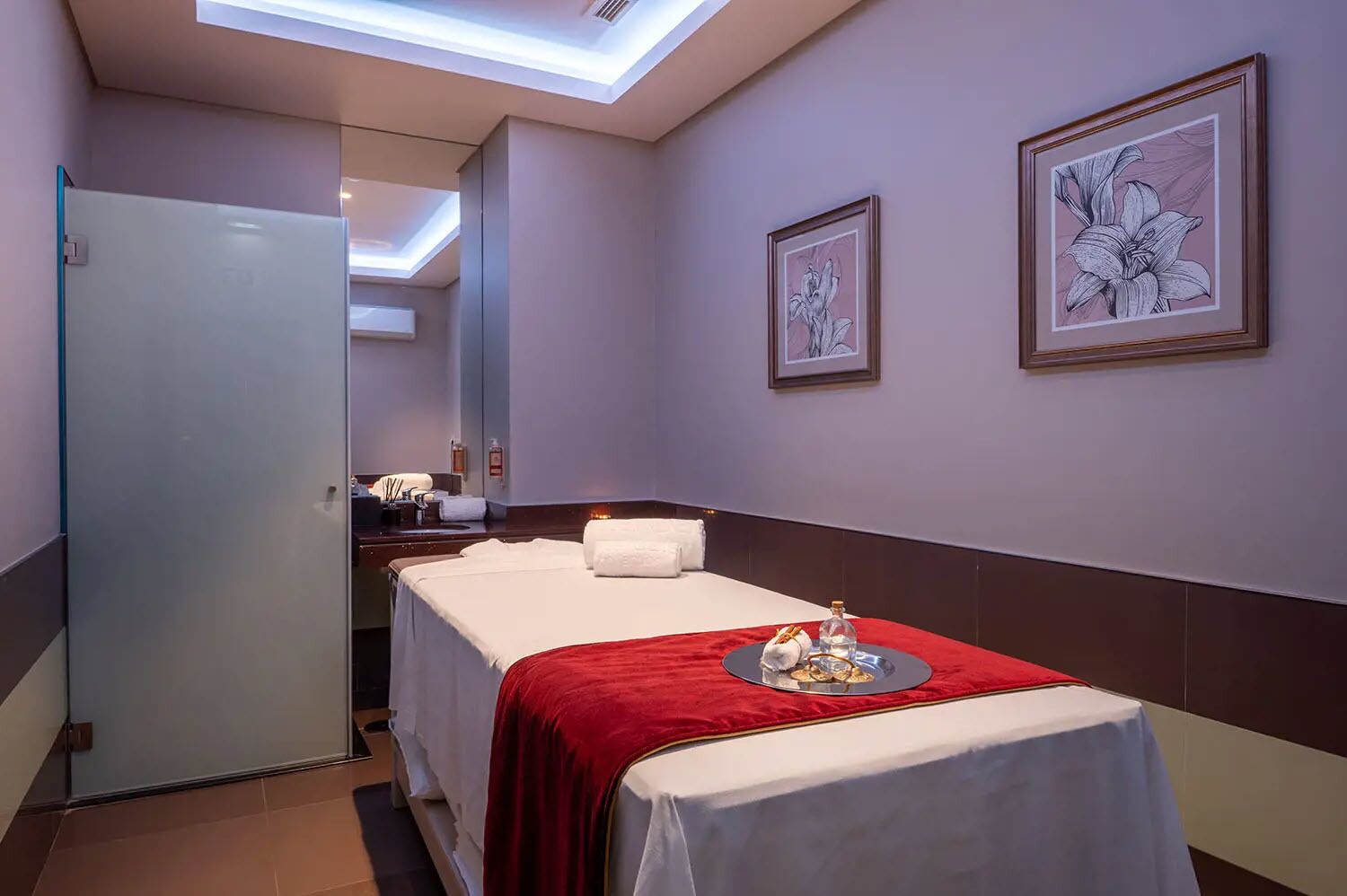 Um espaço de spa com mesa de massagem, uma pequena mesa com uma garrafa e um copo, e duas obras de arte com flores emolduradas na parede.