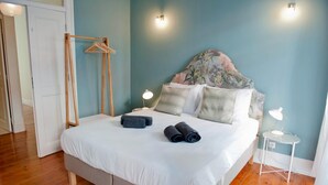 Cama bem arrumada com edredom branco e duas toalhas enroladas, cabide de madeira e mesa de cabeceira pequena com abajur.