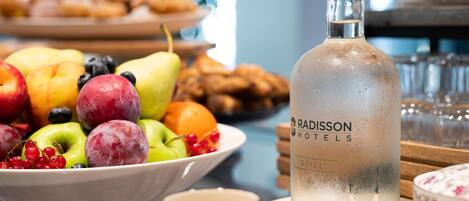 Garrafa de vidro com o rótulo "RADISSON HOTELS", ao lado de uma garrafa de água mineral e uma taça com frutas e doces variados.