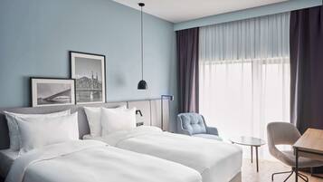 Quarto de hotel com duas camas, uma escrivaninha, uma cadeira e uma janela grande com cortinas.