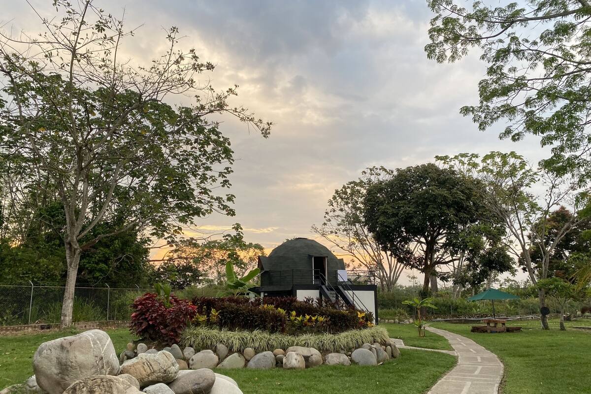Um jardim com um caminho de pedra, uma pequena estrutura em cúpula e árvores ao redor.