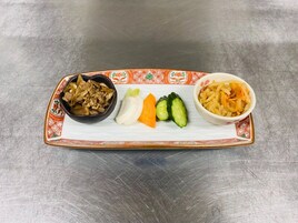 Uma bandeja com diversos alimentos, incluindo um prato com carne, legumes fatiados e um pote de arroz.