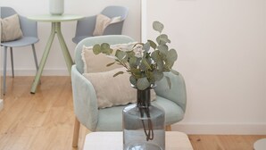 Uma sala de estar moderna com um vaso de vidro transparente sobre uma mesa de madeira, uma poltrona azul clara, uma pequena mesa redonda com uma planta em vaso e uma janela grande com vista para edifícios.
