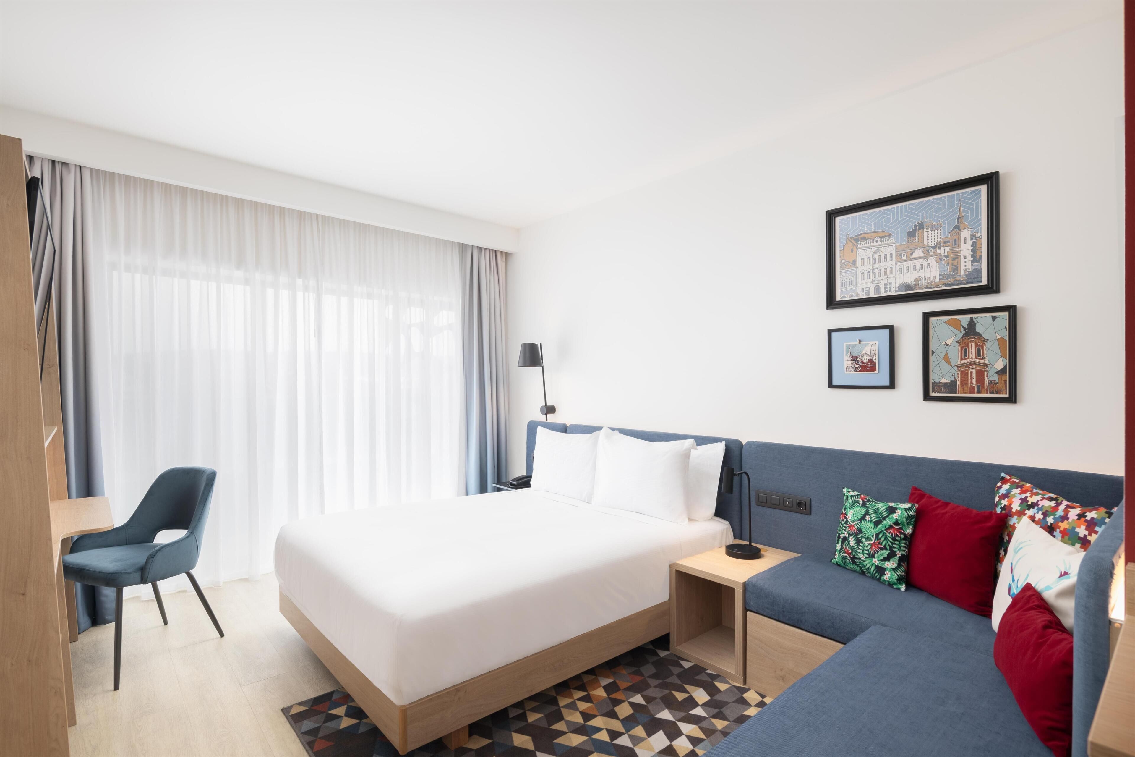 Quarto de hotel com uma cama, escrivaninha, cadeira, sofá e mesa de cabeceira. Três quadros emoldurados na parede.