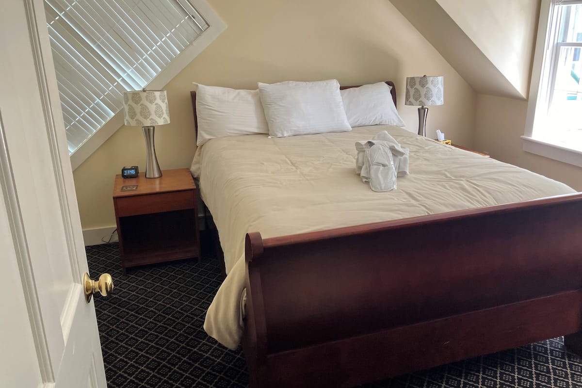 Cama bem arrumada com lençóis e almofadas brancos, um abajur e um relógio em uma mesa de cabeceira de madeira.