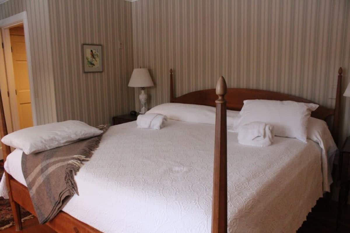 Uma cama bem arrumada com lençóis brancos e cabeceira de madeira, uma mesinha de cabeceira com abajur, e um quadro na parede.