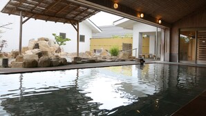 Uma piscina interior calma com um deque de madeira, um recurso de cachoeira de pedra e um edifício em estilo japonês tradicional ao fundo.