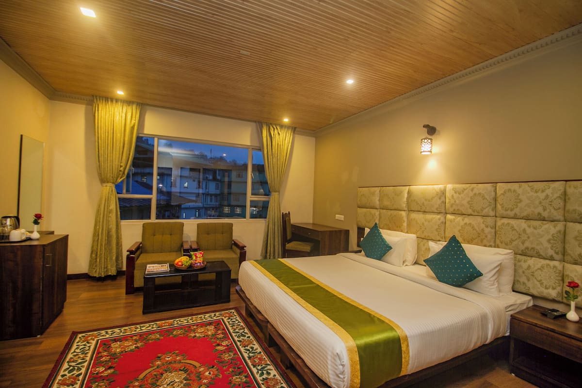 Quarto de hotel com cama grande, escrivaninha, cadeira, janela com cortinas e mesa de cabeceira com um vaso.
