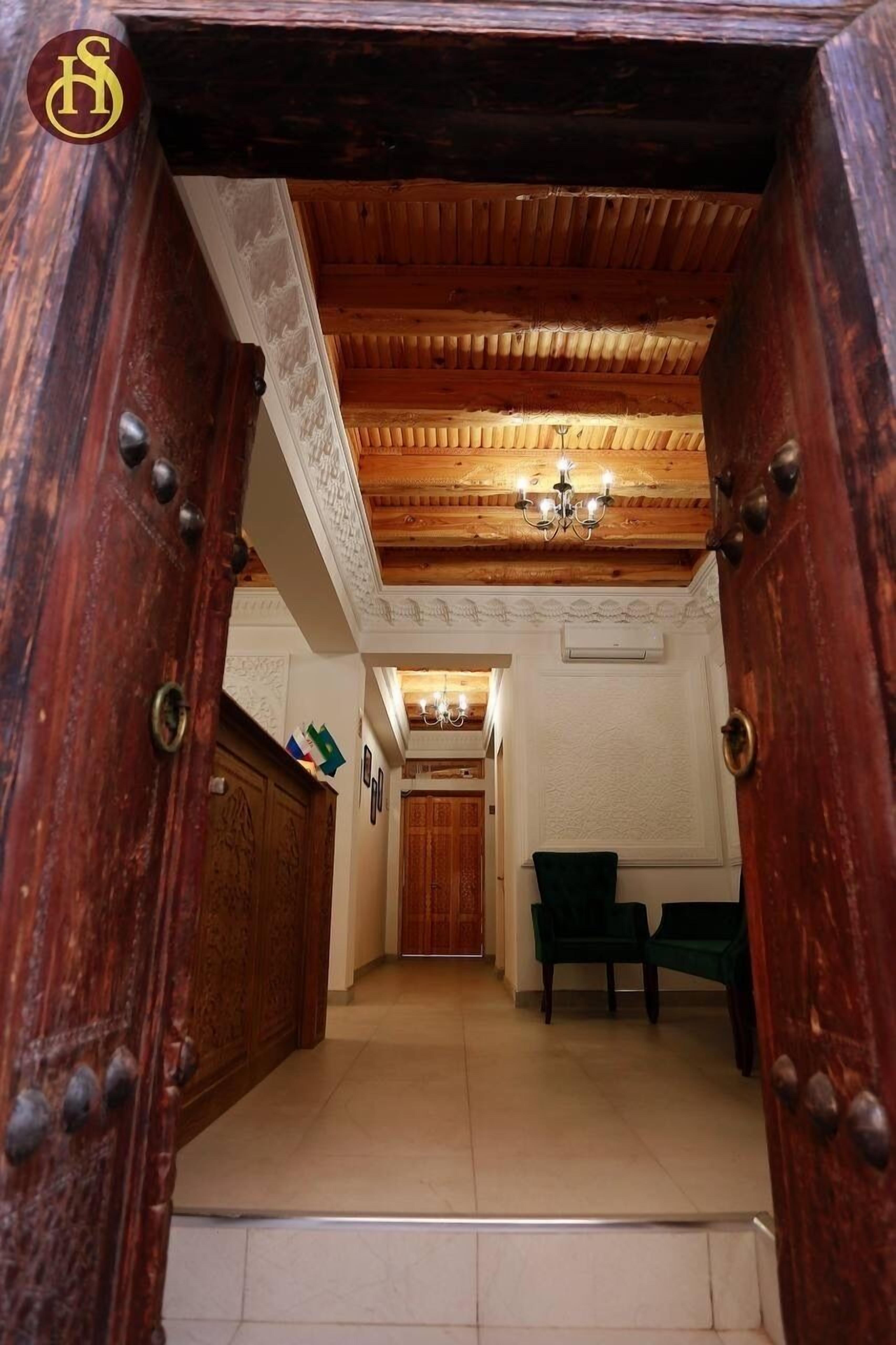 Um corredor de madeira com teto alto, um candelabro e uma porta que dá para um quarto com uma cadeira verde.