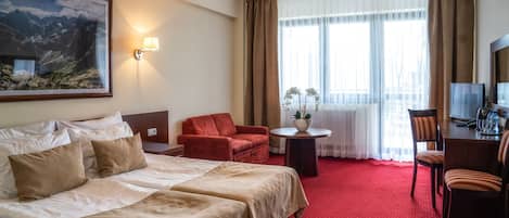 Quarto de hotel com uma cama grande, duas mesas de cabeceira, uma escrivaninha com cadeira, uma pequena mesa com uma planta e uma janela ampla com cortinas.