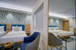 Quarto de hotel com duas camas, uma poltrona azul, uma mesinha com frutas e um ventilador de teto.