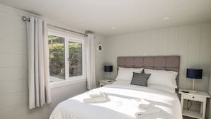 Cama bem arrumada com lençóis brancos, cabeceira, duas mesas de cabeceira com luminárias e uma janela com cortinas.