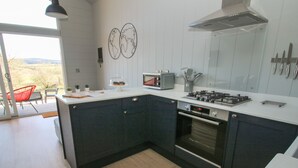 Cozinha moderna com armários escuros, bancada branca, fogão a gás, forno e micro-ondas. Há vista para o exterior por uma porta de correr de vidro.