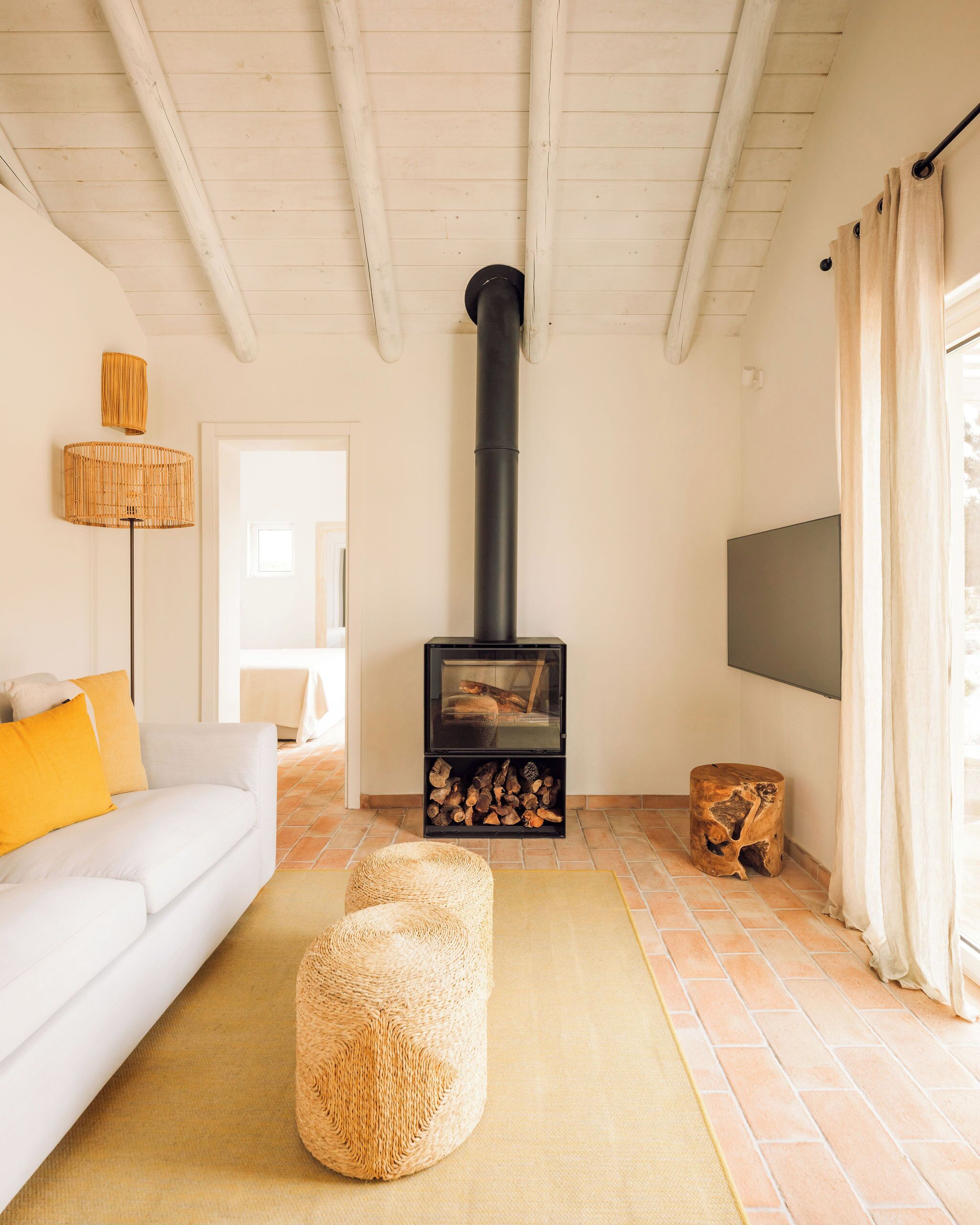 Uma sala de estar com um sofá branco, um banquinho trançado, uma lareira a lenha preta, uma TV e um banquinho de madeira.