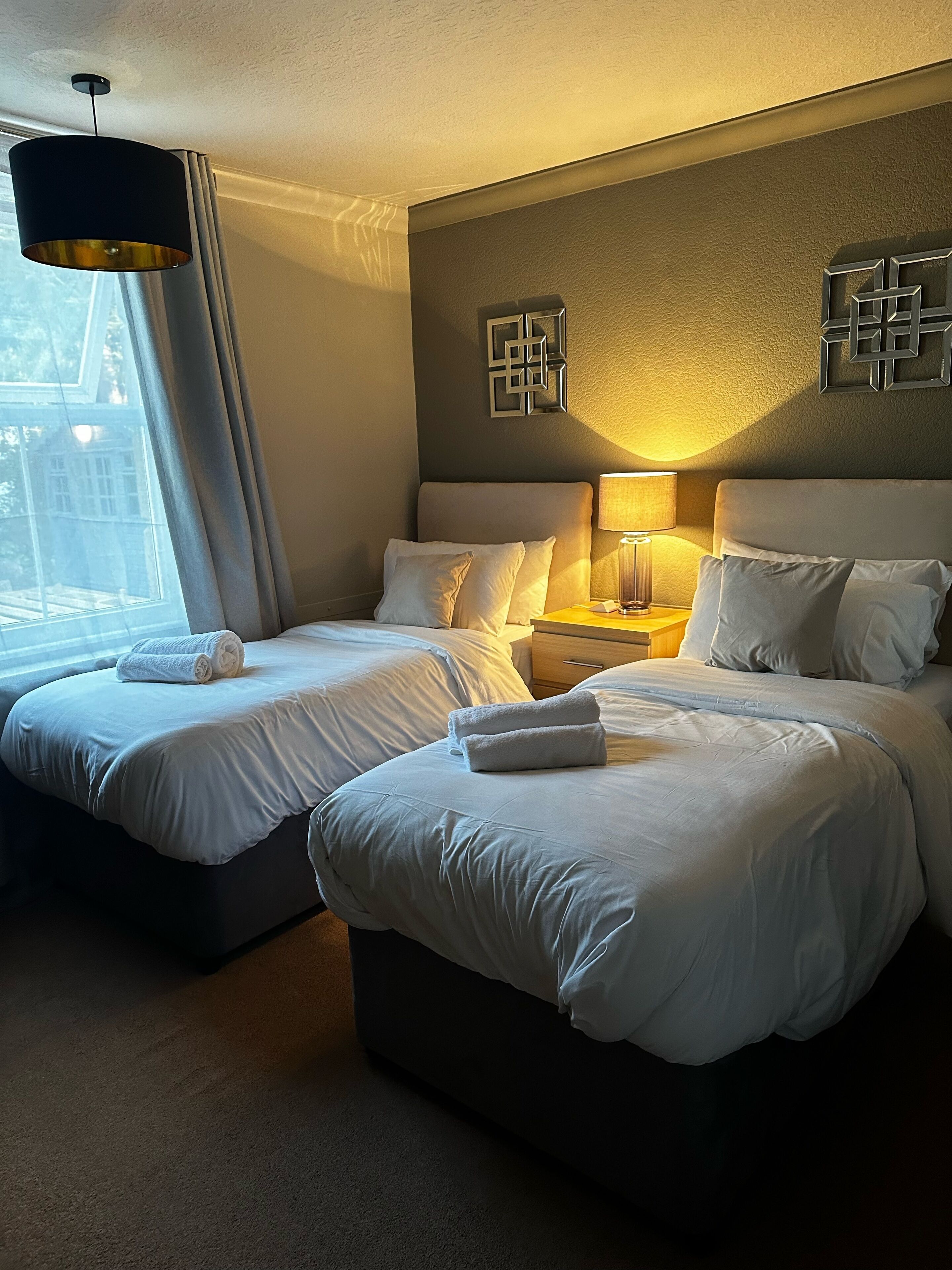 Quarto de hotel com duas camas, uma janela com cortinas, luminária na parede e mesa de cabeceira com abajur.