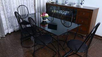 Uma área de refeições com mesa de vidro, quatro cadeiras pretas, um vaso com flores e um armário de madeira com objetos decorativos.