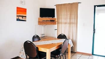 Uma área de refeições com uma mesa de madeira, quatro cadeiras, uma televisão sobre um móvel de madeira, um quadro na parede e uma porta de vidro deslizante.