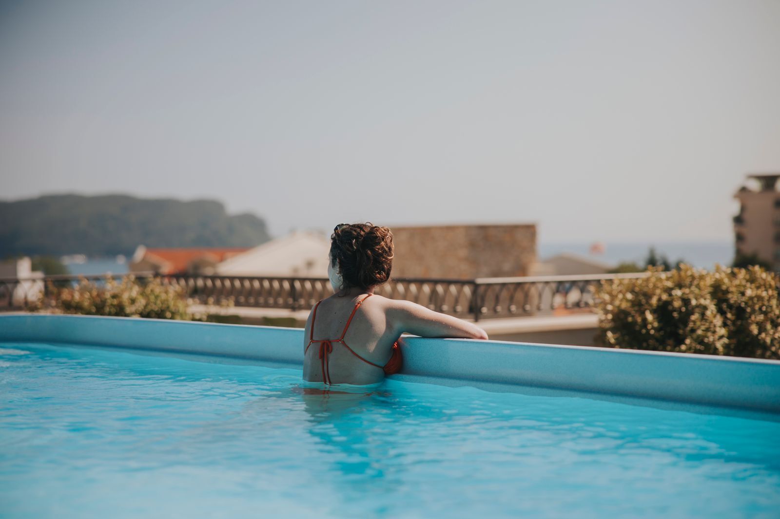 Uma pessoa relaxando em uma piscina com vista para prédios e colinas ao fundo.