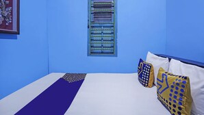 Quarto com uma cama de solteiro, janela com moldura azul e duas almofadas com padrões geométricos.