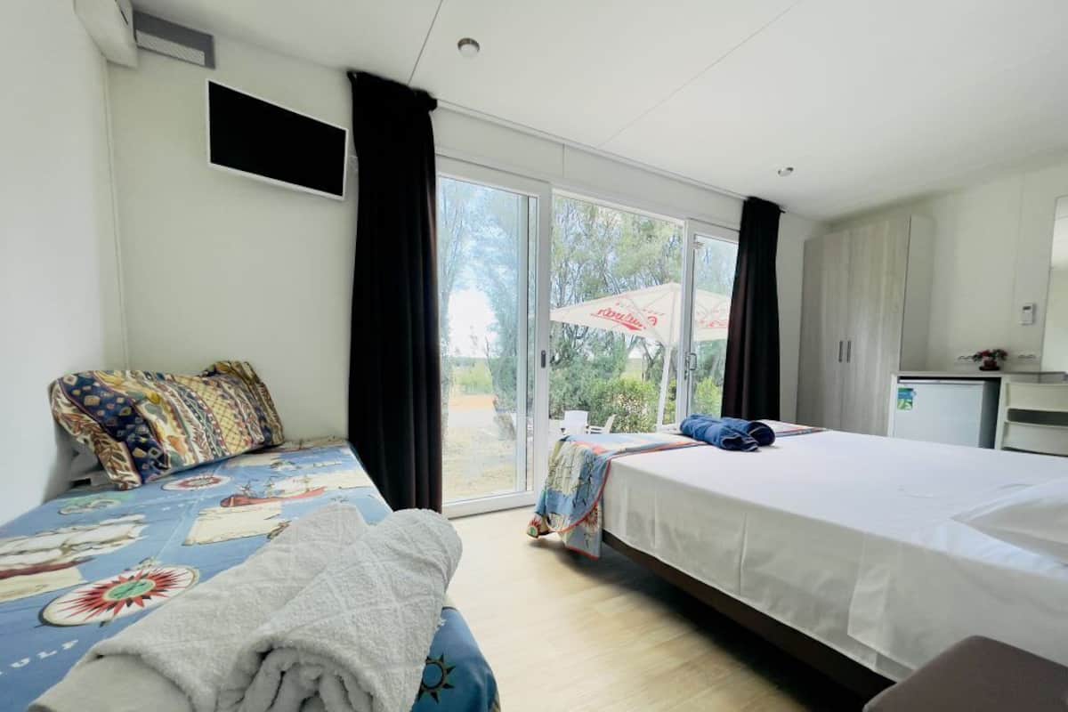 Quarto de hotel com duas camas, televisão, cozinha compacta e porta de vidro deslizante para o exterior.