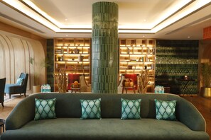 Um lobby moderno com um sofá verde, uma coluna cilíndrica alta e estantes com livros e objetos decorativos.