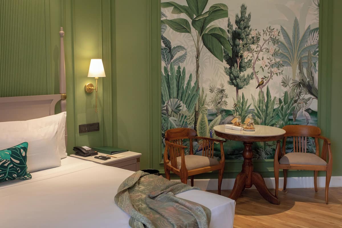 Quarto de hotel com cama, mesa de cabeceira, luminária, mesa com cadeiras e um mural com folhagem tropical.