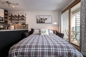 Quarto com colcha xadrez, quadro pendurado de um touro e área de cozinha compacta com prateleiras e uma placa com a inscrição 'BISTRO'.