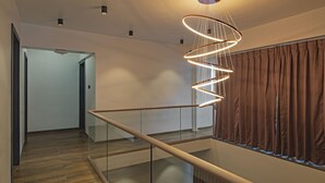 Um interior moderno com guarda-corpo de vidro, luminária em espiral e vista de um quarto através de uma janela com cortinas.