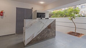 Uma área de recepção com balcão de mármore, uma porta fechada, uma câmera de segurança e uma pequena planta.