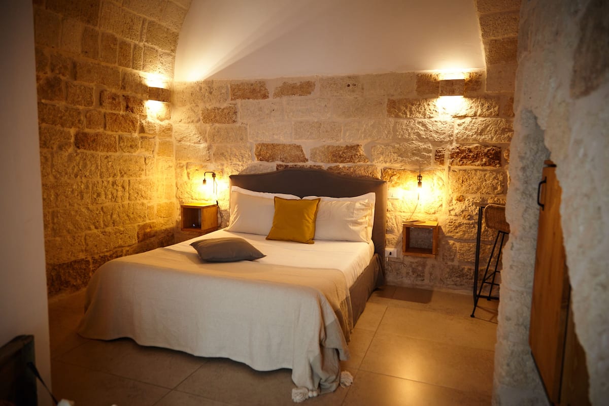 Quarto com parede de pedra, cama com roupa de cama branca e cabeceira escura, duas luminárias de cabeceira e um banquinho de madeira.