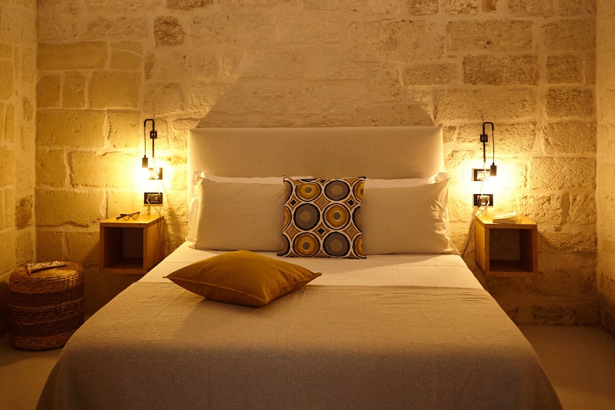 Um quarto com parede de pedra, uma cama com travesseiro estampado e duas luminárias de cabeceira.