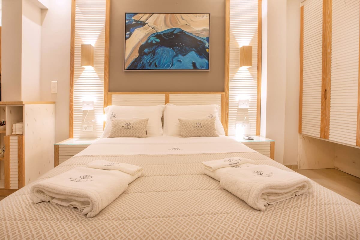 Quarto de hotel com uma cama grande, uma mesa de cabeceira, um criado-mudo, um quadro na parede e uma janela com persianas.
