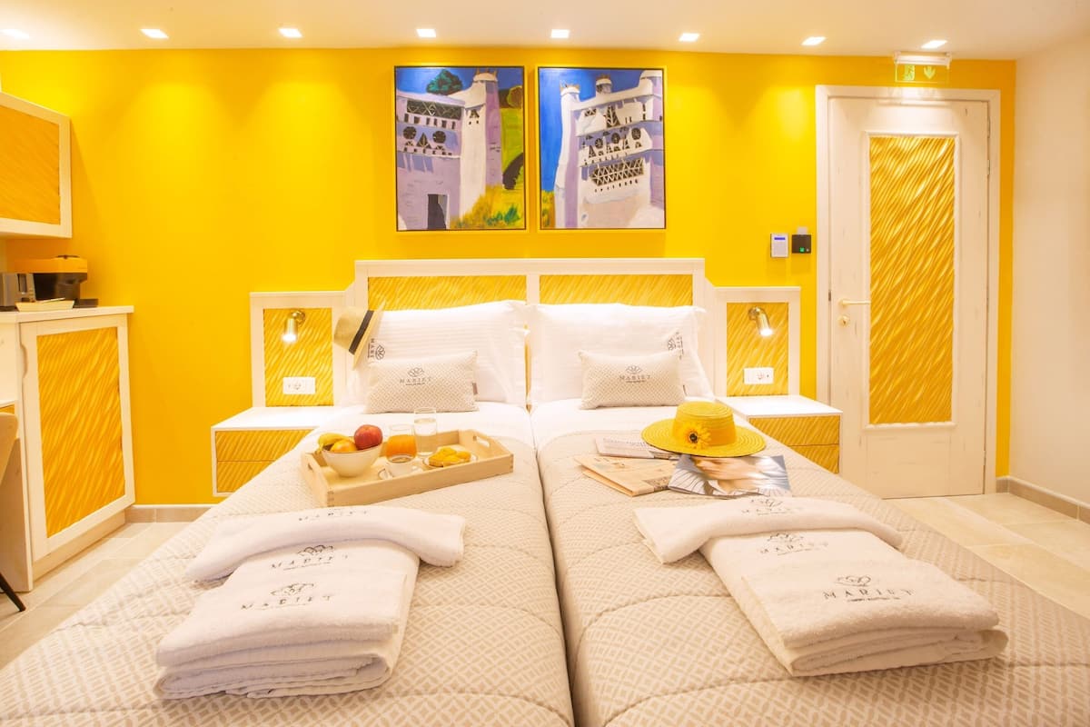 Quarto de hotel com duas camas, uma bandeja de frutas, toalhas e um chapéu sobre a cama.