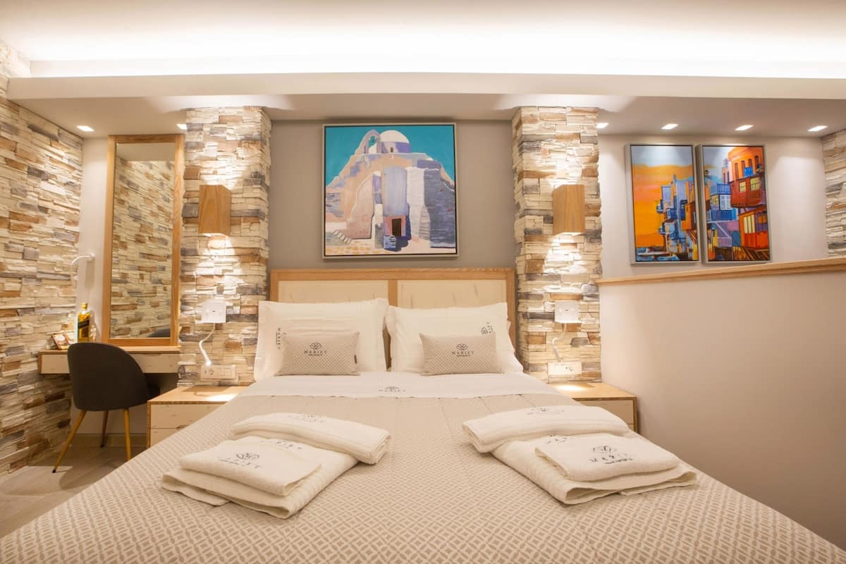 Quarto de hotel com uma cama grande, duas mesas de cabeceira, uma escrivaninha e uma cadeira. Há duas pinturas na parede, um espelho e uma parede de pedra.