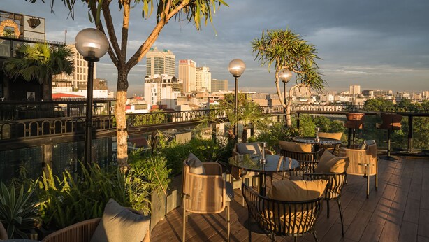 Terraço no telhado com área de estar externa, palmeiras e horizonte urbano ao fundo.